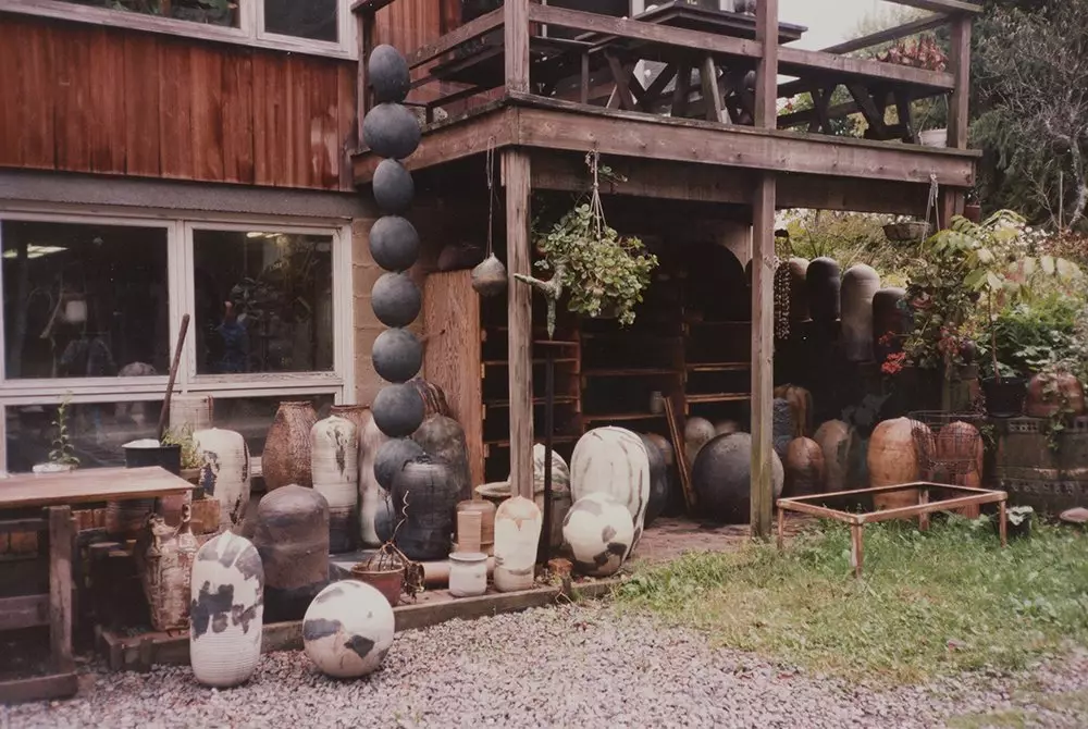 Colección de grandes vasos de cerámica, na súa maioría en marróns e tons terra, fóra dunha casa de madeira.