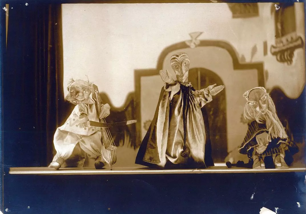 Три куклы ручной работы стоят перед декорацией дворца. Марионетка слева держит опорную виолончель, марионетка посередине стоит с распростертыми руками, а марионетка справа присела.