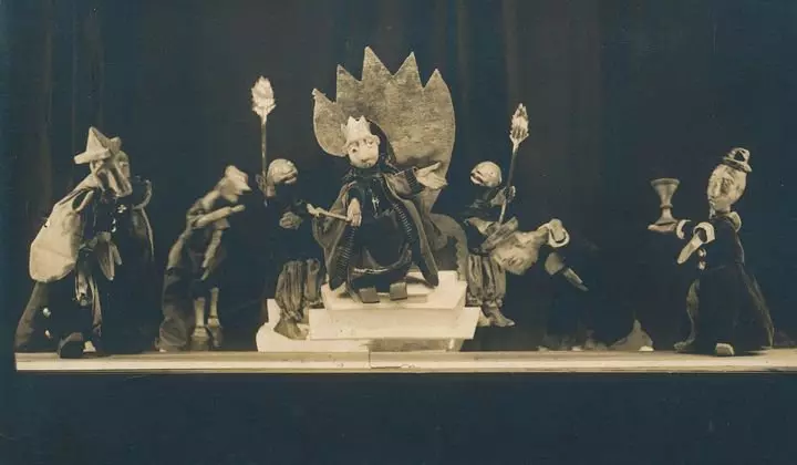 Несколько марионеток на сцене, в центре — король на троне. Черно-белое фото.
