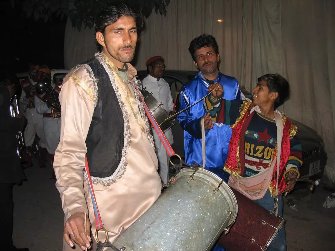 A nighttime wedding procession. A man in front carries a metal barrel-shaped drum over his shoulder and glares at the camera. Others in the background carry drums and brass instruments.