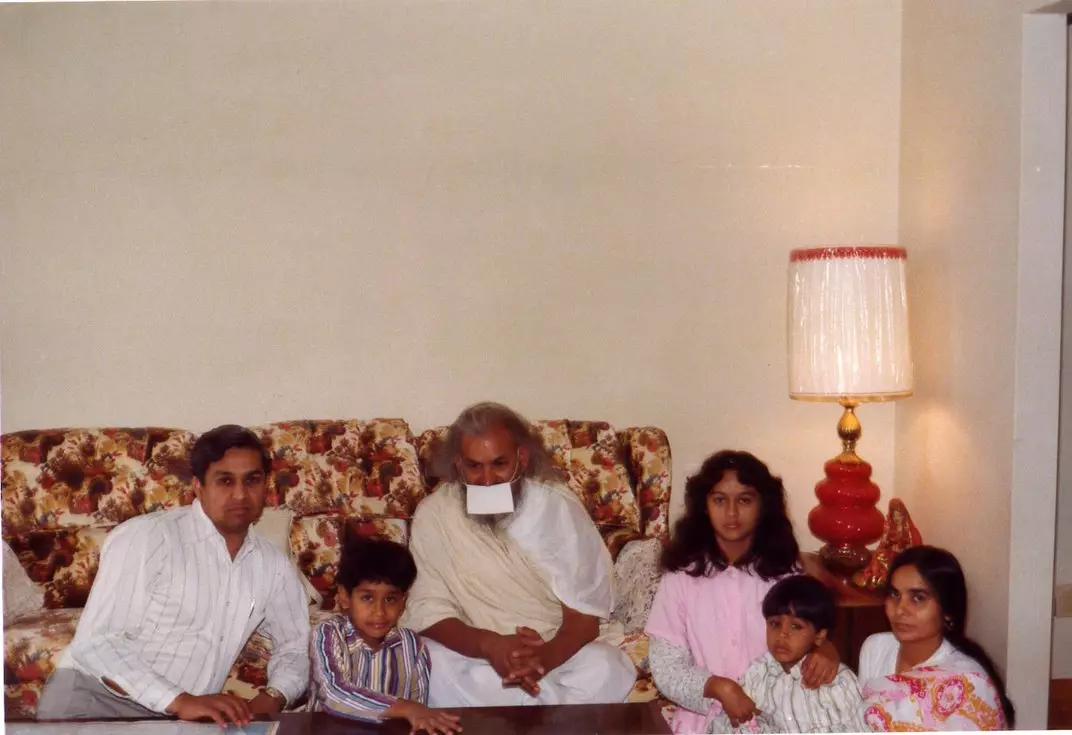Old family photo with the Jain family and an older man posing, sitting on and around a beige floral-patterned couch. The older man is wearing a white face mask.