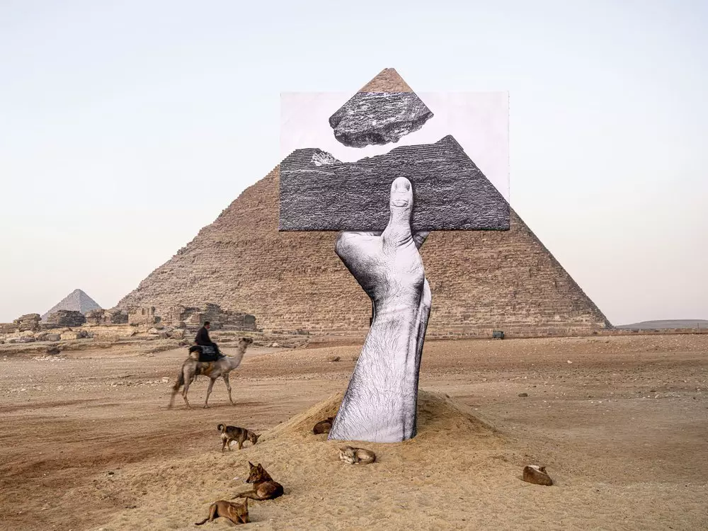 En ulegemelig sort og hvid hånd kommer frem fra sandet og holder et fotografi, hvilket skaber den effekt, at spidsen af en pyramide bagved er adskilt og svæver over dens base