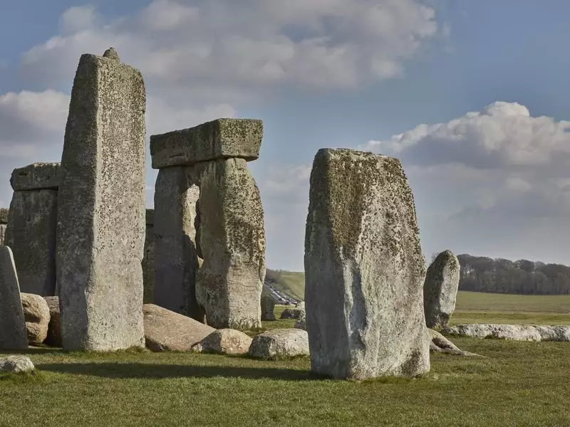 Widok na kultowe skały Stonehenge na zielonej trawie, z pochyłym wzgórzem widocznym między dwoma kamieniami w oddali i pasem ruchu