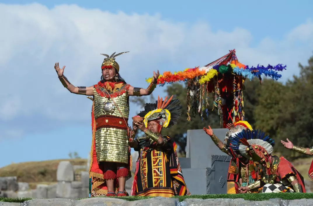 Een man staat met opgeheven armen, gekleed in een glanzende gouden outfit, een gouden gevederde pet en een gouden borstplaat. Daarachter regenboogkleurige veren en twee mensen die knielen en hun handen naar hem opheffen