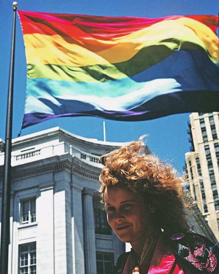 In frou mei brún krolhier stiet en glimket foar in reinbôgekleurige flagge dy't yn 'e wyn waait foar in statich wyt gebou en blauwe loft