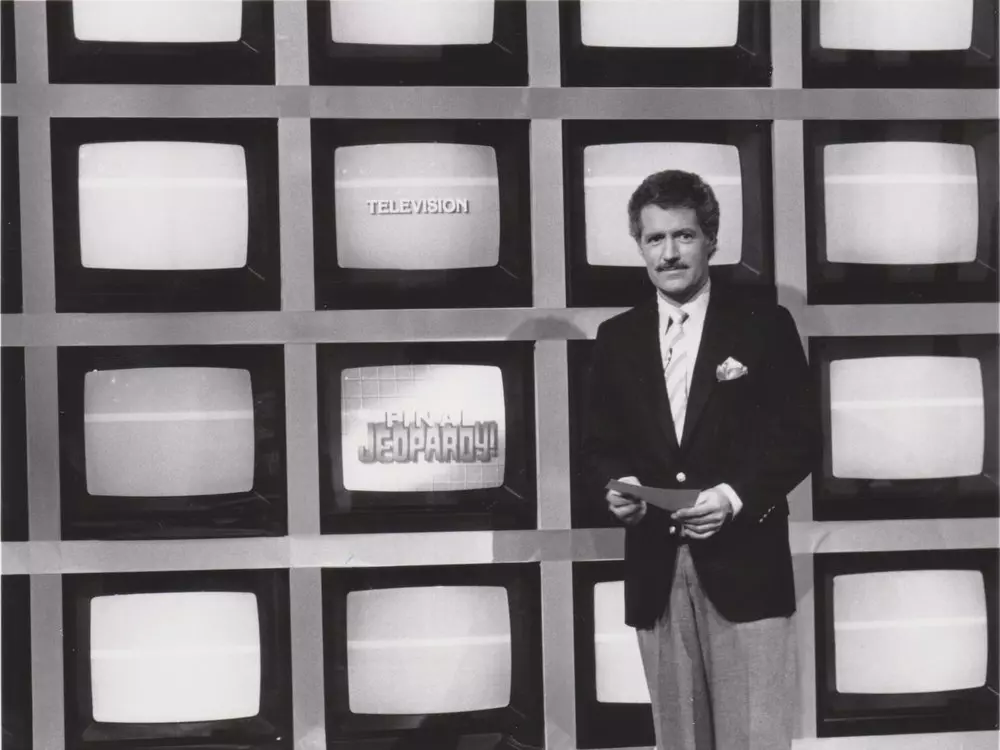 ภาพขาวดำของหนุ่ม Trebek ชายผิวขาวในชุดสูทและผูกผมสีเข้มและหนวด ยืนอยู่หน้าตารางทีวี