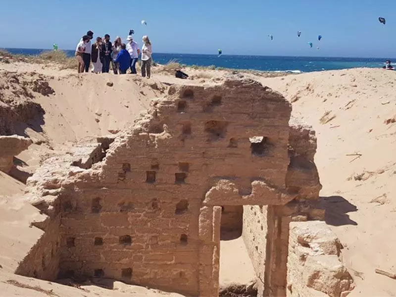 Een groep mensen staat voor een blauwe lucht en een blauwe zee terwijl ze naar een structuur kijken, met veel kleine gaten zoals ramen en hoge muren