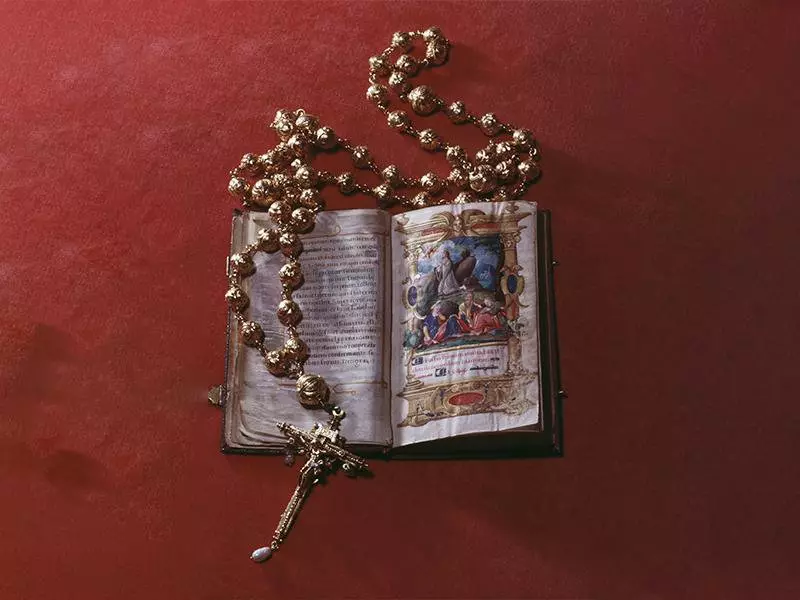 在红色背景上，一本打开的照明圣经，上面有彩色插图，描绘了耶稣跪下和祈祷，周围环绕着一个带有大圆珠的金色念珠和一个饰有珍珠的十字架，以及一个被钉在十字架上的耶稣的金色形象