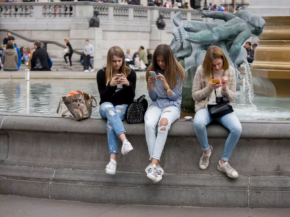 Три тинејџера седе са стране велике фонтане и гледају у своје паметне телефоне
