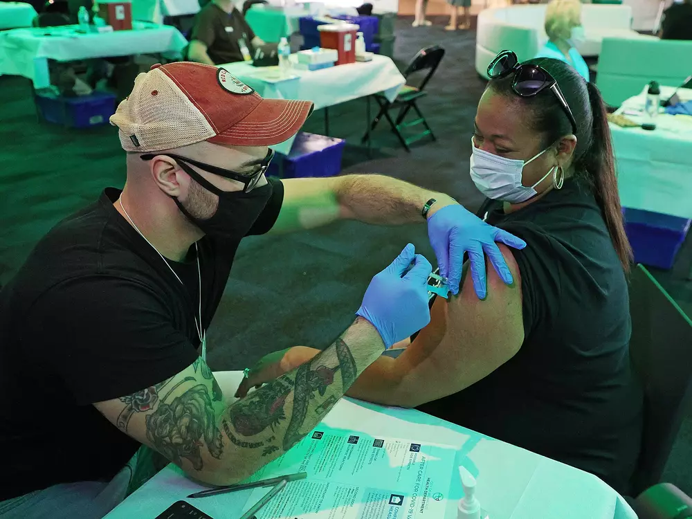 Žena v čiernej košeli a slnečných okuliaroch dostane vakcínu od muža v čiernej košeli a červenej baseballovej čiapke, obaja sedia pri bielom stole a majú masky