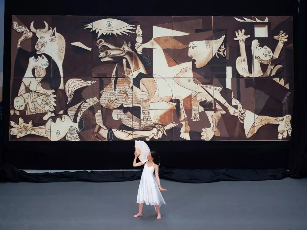 A person in a white flowy dress and holding a white fan dances in front of the enormous chocolate replica of Picasso's Guernica