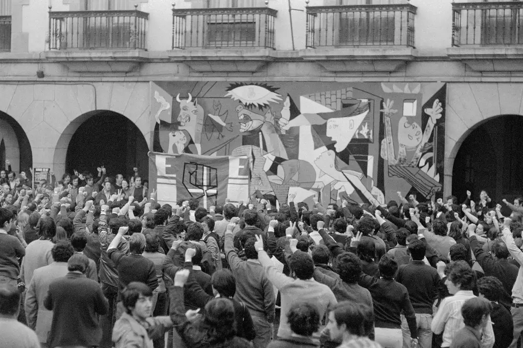 A black and white image of a crowd of young people, all raising their fists and looking at a copy of Picasso's Guernica hung from a large balcony