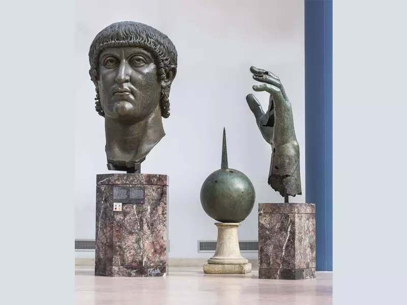 A view of a huge bronze cast head of a man with tightly curled hair and a serious expression, a sphere and his hand, all fragments that once were part of a huge 12m-tall sculpture