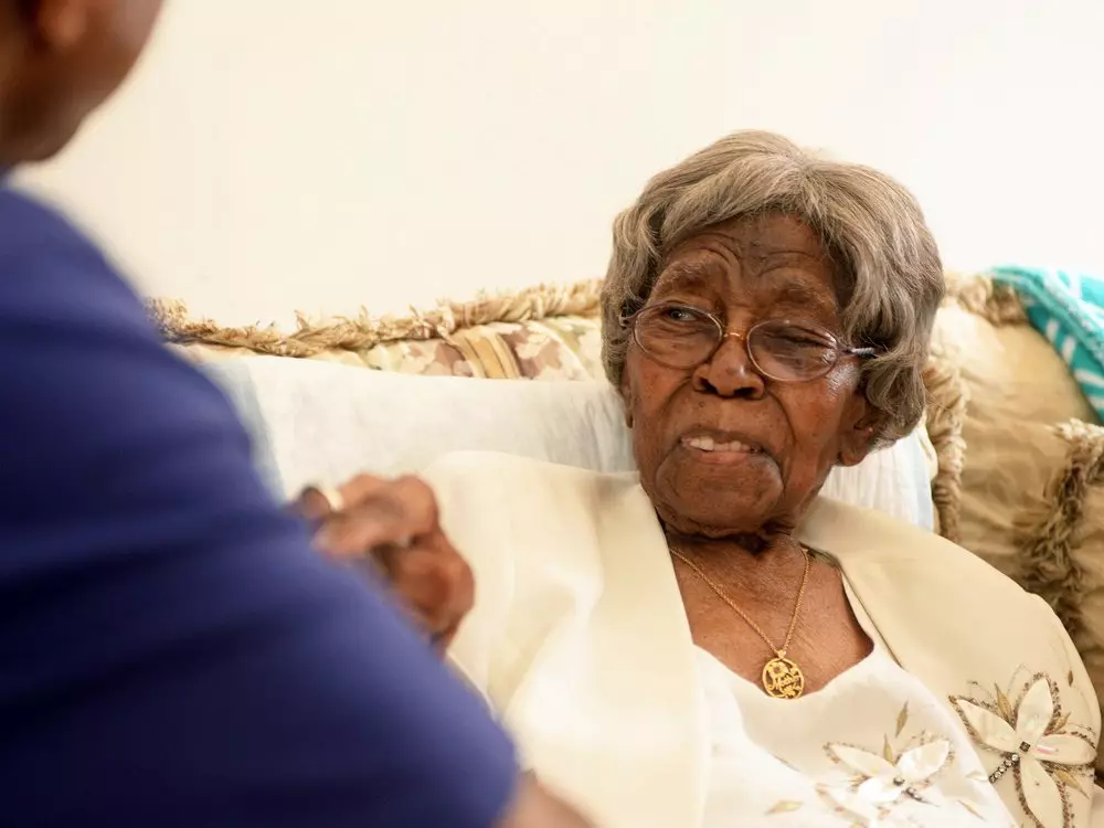 Unha muller negra de idade avanzada, con cabelos curtos e grises, sentada nun sofá e sorrí detrás das lentes, leva un colar de ouro e un xersei branco.