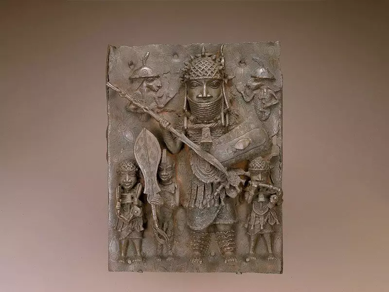 Plak logam coklat yang diukir dengan figura, termasuk figura tengah besar yang memakai perisai dan menjulang tinggi di atas sekurang-kurangnya tiga orang yang lebih kecil berdiri di bawah
