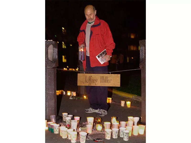 Een man in een rood jasje kijkt neer op kaarsen rond een bord met de tekst 'Jeffrey Miller'