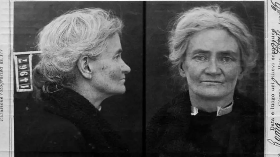 Une photo criminelle en noir et blanc d'une femme blanche aux cheveux grisonnants, dans une tenue noire