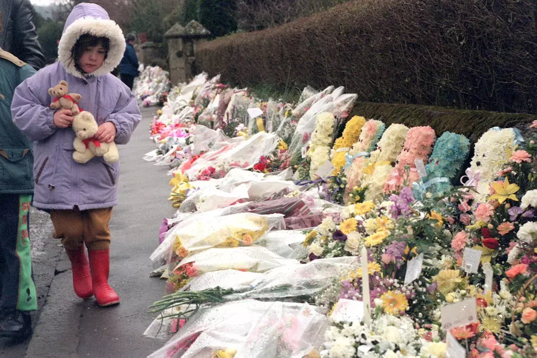 एक जवान लड़की अपने मारे गए सहपाठियों और शिक्षक का सम्मान करते हुए फूलों की कतार से गुजरती है