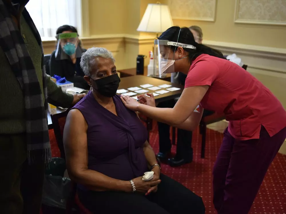 Një grua ulet në një karrige dhe merr një vaksinë nga një grua e veshur me maskë dhe mburojë për fytyrën