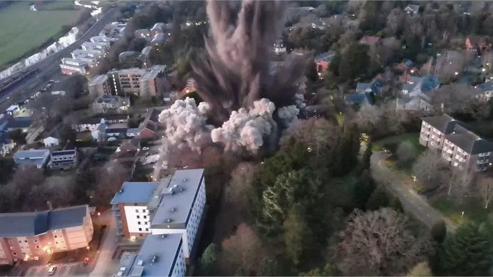 Una vista aérea de una gran explosión que tiene lugar en medio de un vecindario, con nubes de polvo y humo que emergen de los edificios y están rodeadas de árboles verdes.