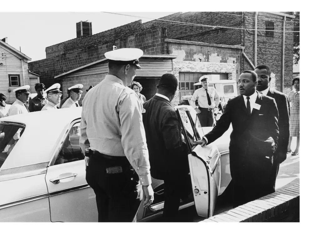 Uma fotografia em preto e branco do policial em primeiro plano, um homem entrando em uma viatura e MLK com um braço estendido movendo-se em direção ao carro