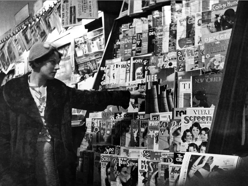 ایک نوجوان عورت کی سیاہ اور سفید تصویر، ٹوپی اور فر کوٹ پہنے ہوئے، ایک پرہجوم نیوز اسٹینڈ پر لائف کی کاپی کے لیے پہنچ رہی ہے۔ نیویارکر اور ٹائم کی ایک کاپی بھی نظر میں ہے۔