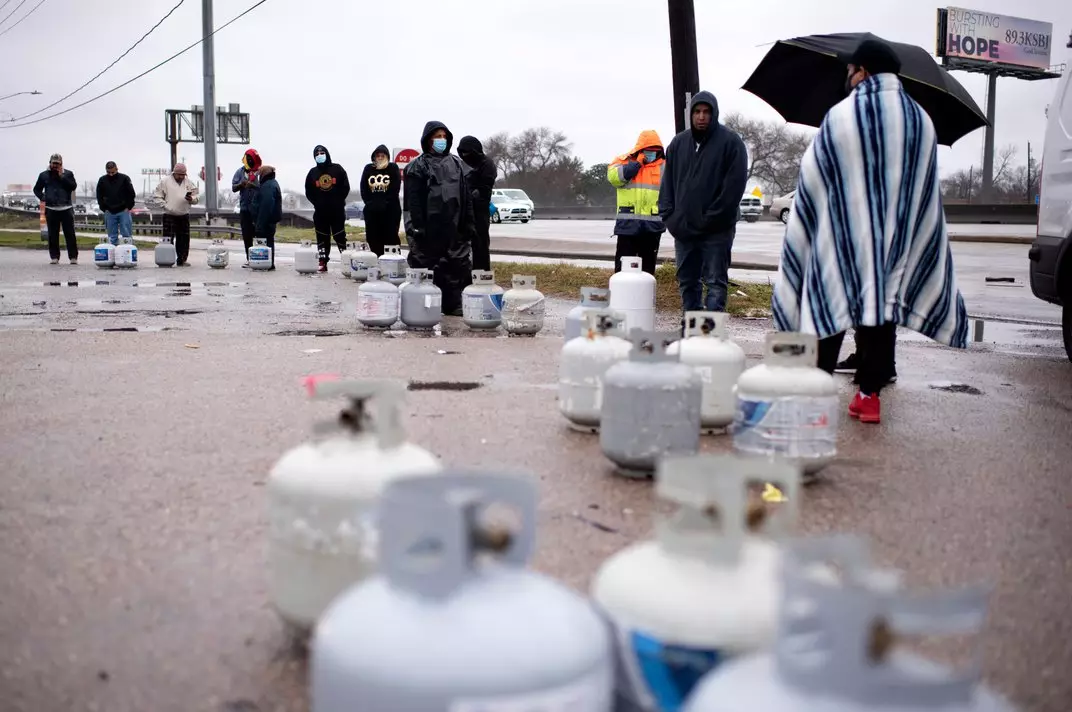 Une ligne de personnes debout à côté de réservoirs de propane sur l'asphalte avec des flaques d'eau