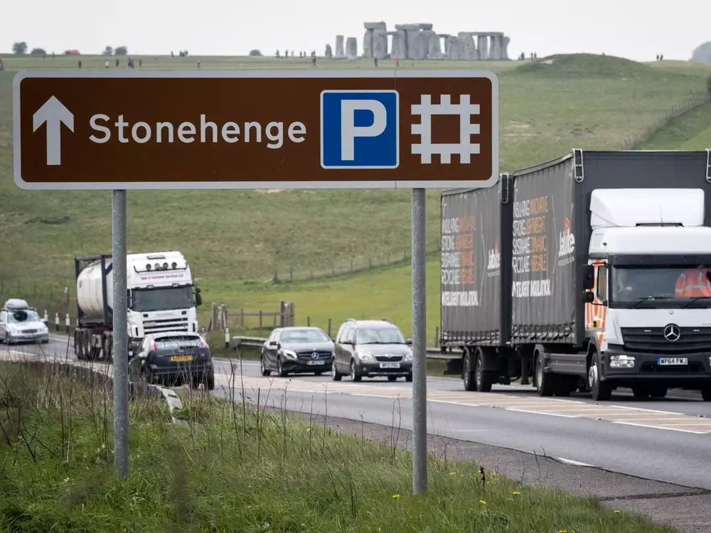 In drokke dyk is op 'e foargrûn, mei frachtweinen en auto's, en in grut teken dat nei Stonehenge wiist; de byldbepalende stiennen struktueren binne sichtber op de eftergrûn