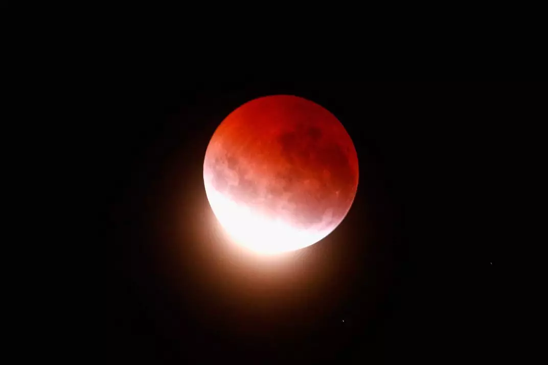 Una lluna gairebé completament eclipsada, amb llum blanca brillant a la seva corba inferior esquerra i ombra majoritàriament vermellosa que cobreix la resta de la seva superfície
