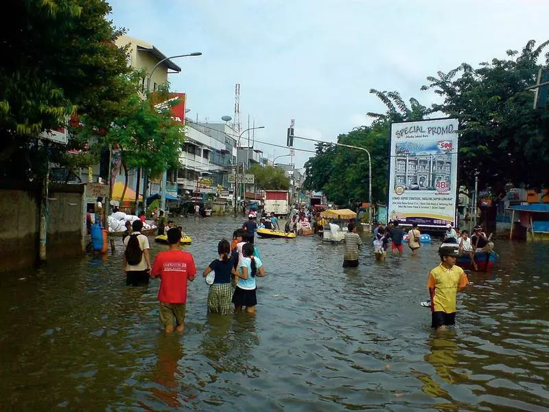 Изглед към наводнен пазар в Джакарта. Хората газят през вода до колене насред улица. Изглежда като типична, оживена улица, с изключение на това, че улицата е изцяло наводнена.