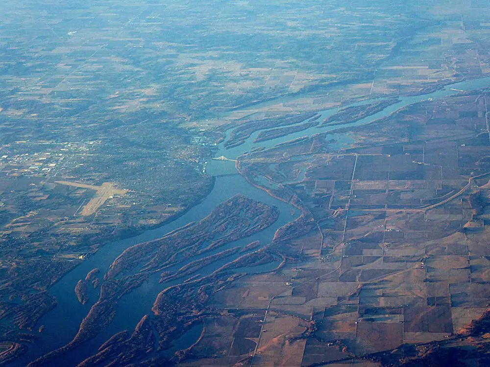 Una foto aérea del río Mississippi que fluye a través de Iowa. El río corre por el centro de la foto, separando parches de campos a la derecha y áreas más urbanas y suburbanas a la izquierda.