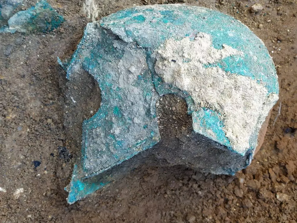 Vue d'un casque en métal oxydé bleu-vert, avec des plaques de joue incurvées pointues qui s'étendent au-delà du visage, allongé sur le sol dans la saleté