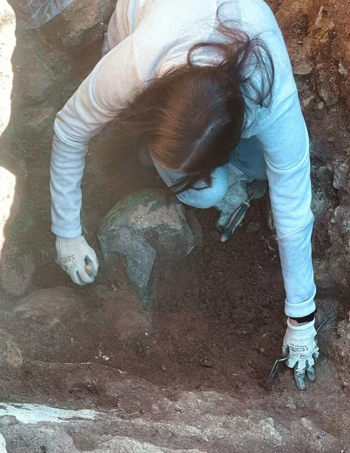 En arkeolog bøyer seg over hjelmen, som er halvt utgravd og ser grønn ut på grunn av oksidasjon