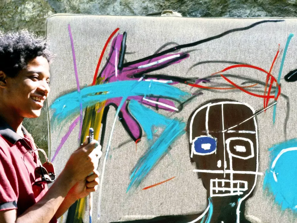 Jean-Michel Basquiat, brosandi, stendur fyrir framan eitt af verkum sínum í vinnslu. Það er sólskin úti og hann er í rauðri skyrtu