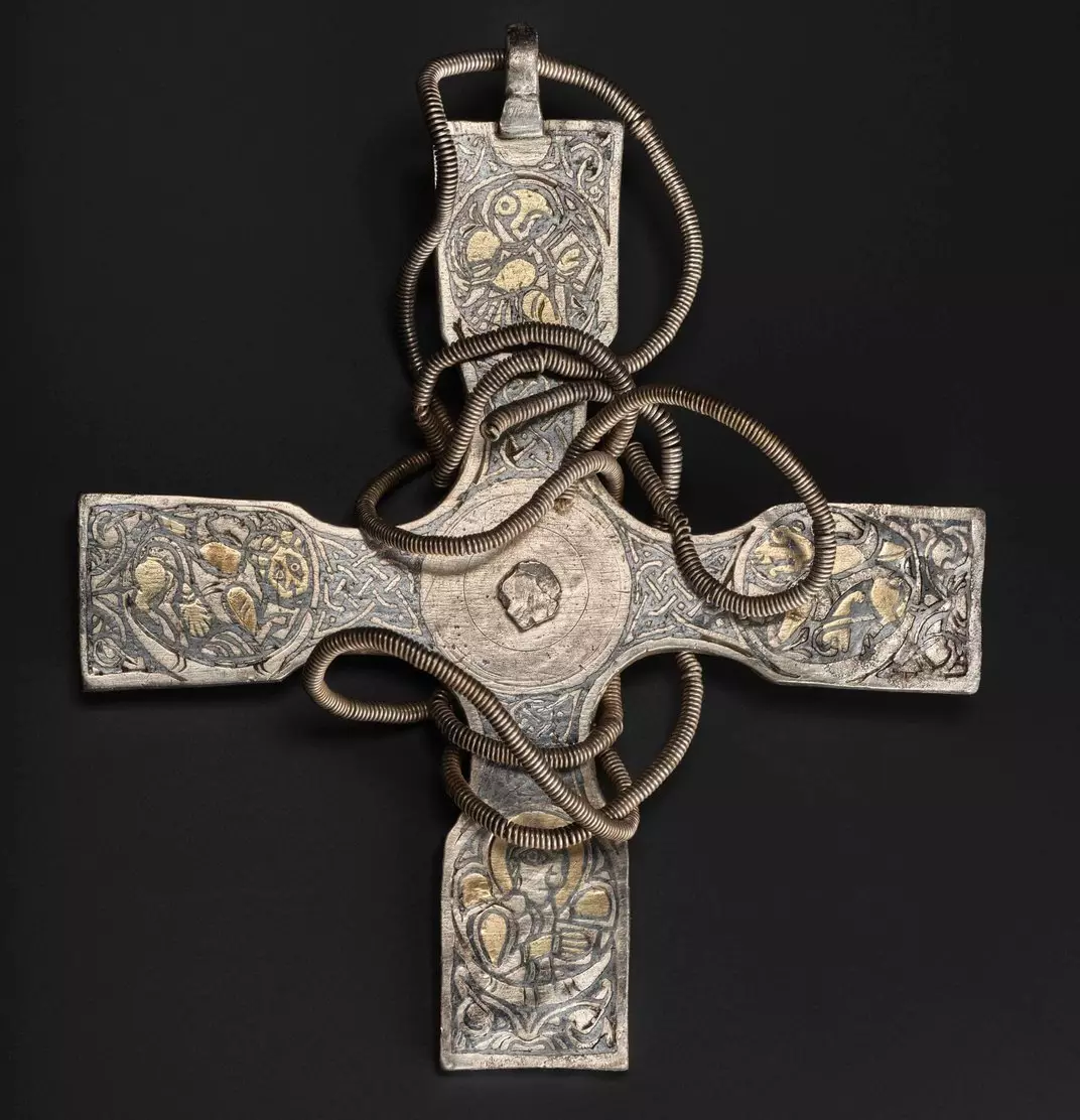 Uma cruz de prata, com um fio que se enrola em torno de sua superfície e quatro braços igualmente longos gravados com delicadas representações de um humano, vaca, leão e águia