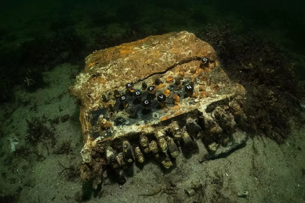 Un primo piano della macchina arrugginita sul fondo dell'oceano, un po' ricoperta di alghe arancioni ma con i suoi tasti, come quelli di una macchina da scrivere, ancora distinguibili