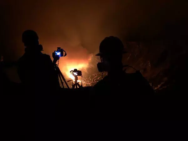 تصویری از فوران آتشفشان. دو نفر با دوربین در پیشزمینه ایستادهاند، اما فقط سیلوئتهایشان قابل مشاهده است. در پسزمینه، یک ابر نارنجی از بخار بالا میآید و زمین دارای ترک نارنجی روشن است.