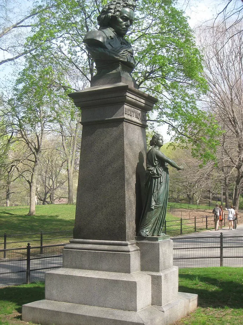 Ein von viel Grün umgebener Sockel im Central Park, auf dem Beethovens Büste nach unten schaut und eine kleinere Figur einer Frau in Roben unter ihm steht