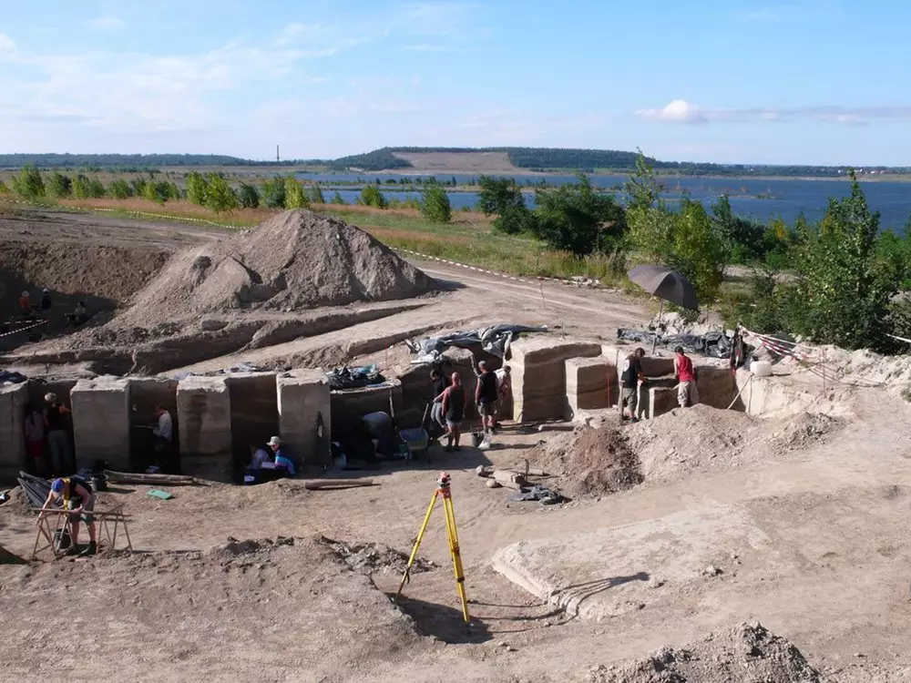 Изображение на археолози при разкопки на обект. Те стоят в яма близо до купчина пръст. В околността има езера и дървета.