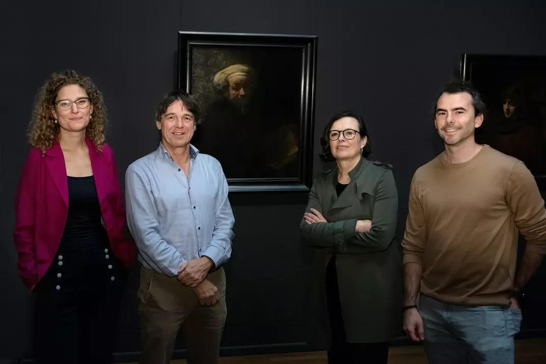 Cuatro personas junto al retrato de un anciano en una galería oscura
