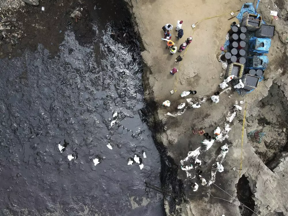 Isang aerial na imahe ng isang oil spill sa baybayin ng Peru. Ang larawan ay nagpapakita ng isang kontaminadong beach na pinahiran ng itim na krudo at mga manggagawa sa PPE na sinusubukang linisin ang beach.