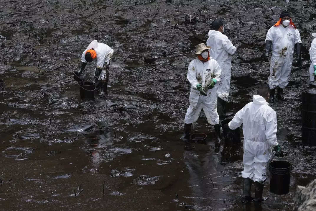 Gambar kru pembersihan mengenakan jumpsuits putih dan sepatu bot karet berdiri di lumpur minyak. Beberapa pekerja memegang ember.