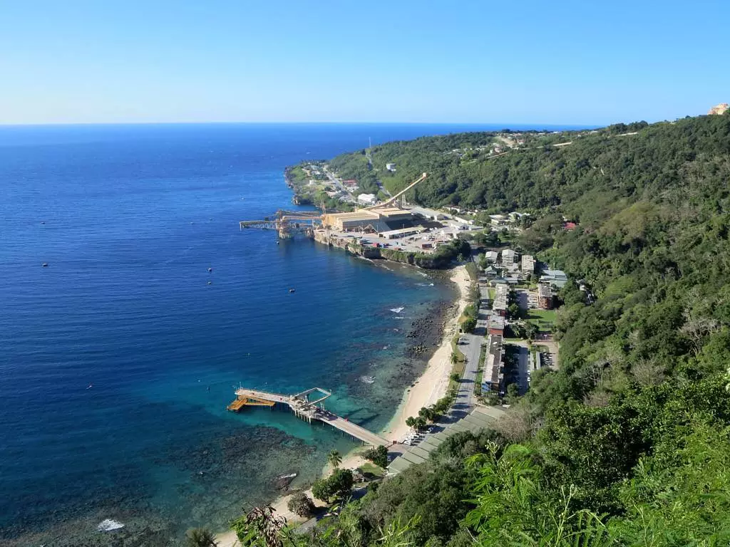 Uma imagem de uma cidade na costa da Ilha Christmas. O lado direito da foto tem colinas arborizadas com uma cidade estendida ao longo da costa. À esquerda está o oceano azul brilhante.